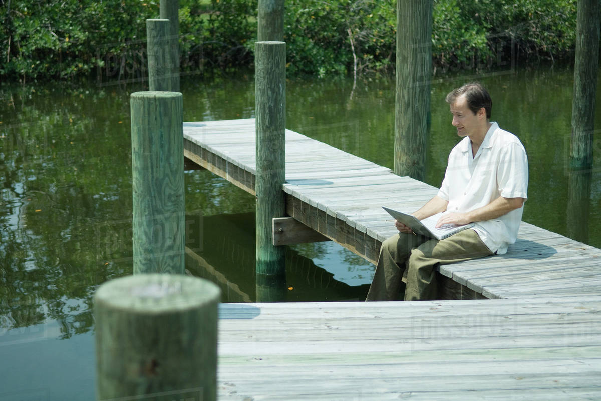 Man sitting on dock, using laptop computer, looking away, smiling ...