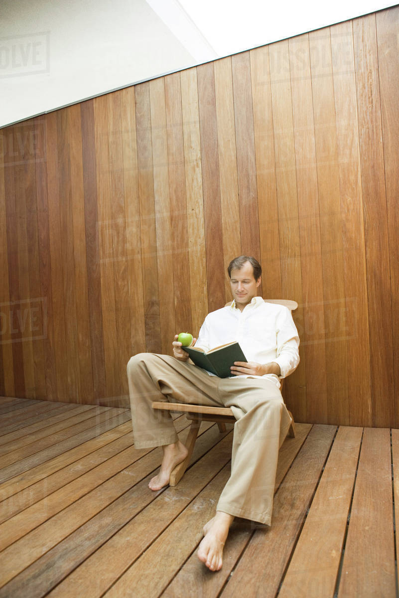 Man sitting in lounge chair reading book, apple in hand - Royalty-free ...