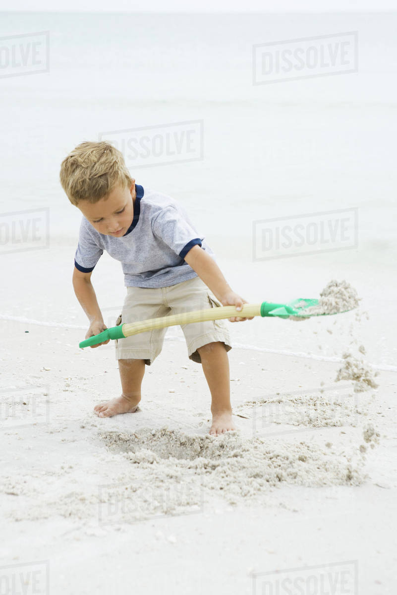 Young boy standing on beach, digging in sand with shovel, looking down ...