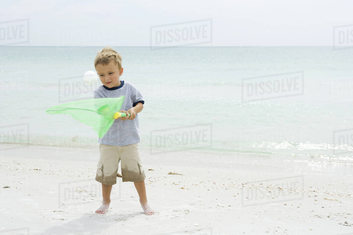 Young boy standing at the beach, trying to catch ball with net ...