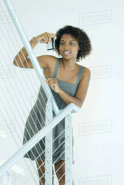 Young woman leaning against railing, holding up digital camera ...