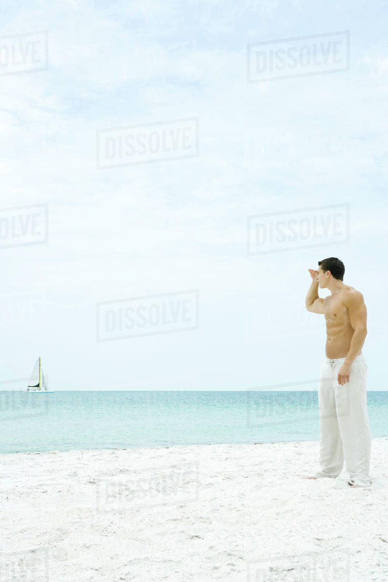 Man standing at the beach, looking at sailboat in the distance, side ...
