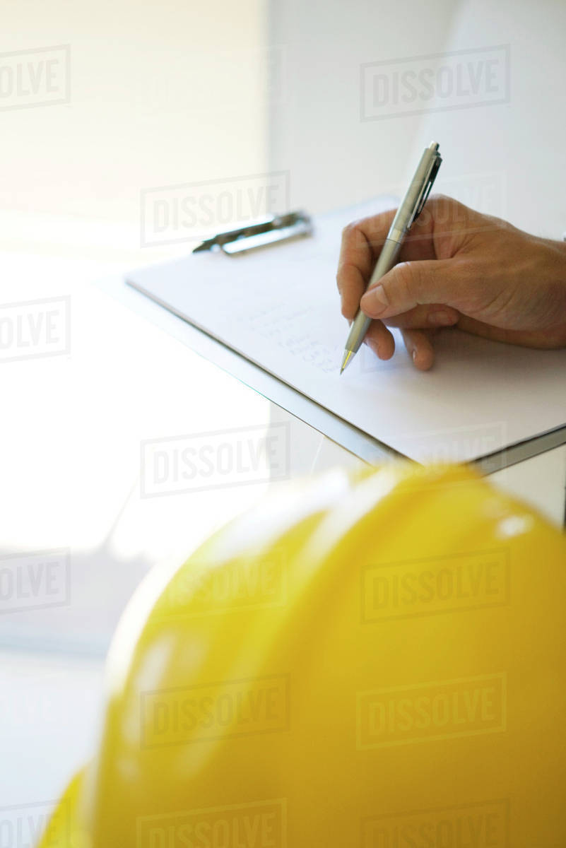 Hand writing notes on clipboard pad, hard hat in foreground, cropped ...