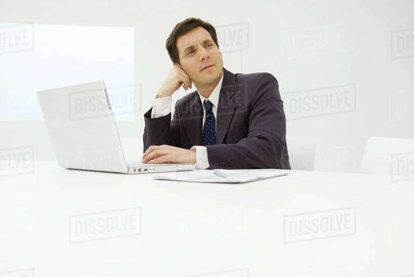 Businessman using laptop computer at desk, holding head, looking away ...