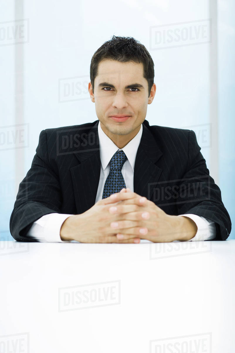 Businessman sitting at table with hands clasped, looking at camera ...