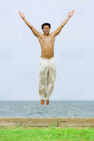 Man jumping in the air with arms raised, smiling at camera, the sea in ...