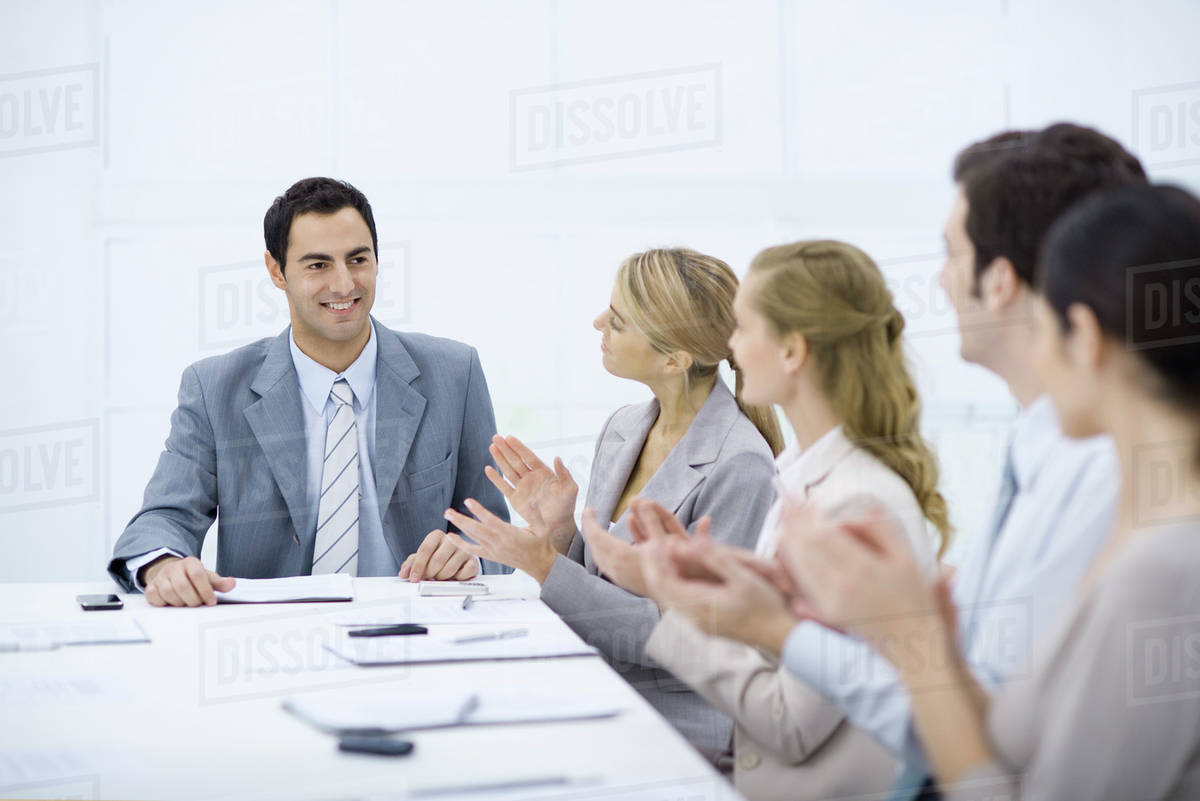 Businessman seated at head of table, smiling, colleagues clapping ...