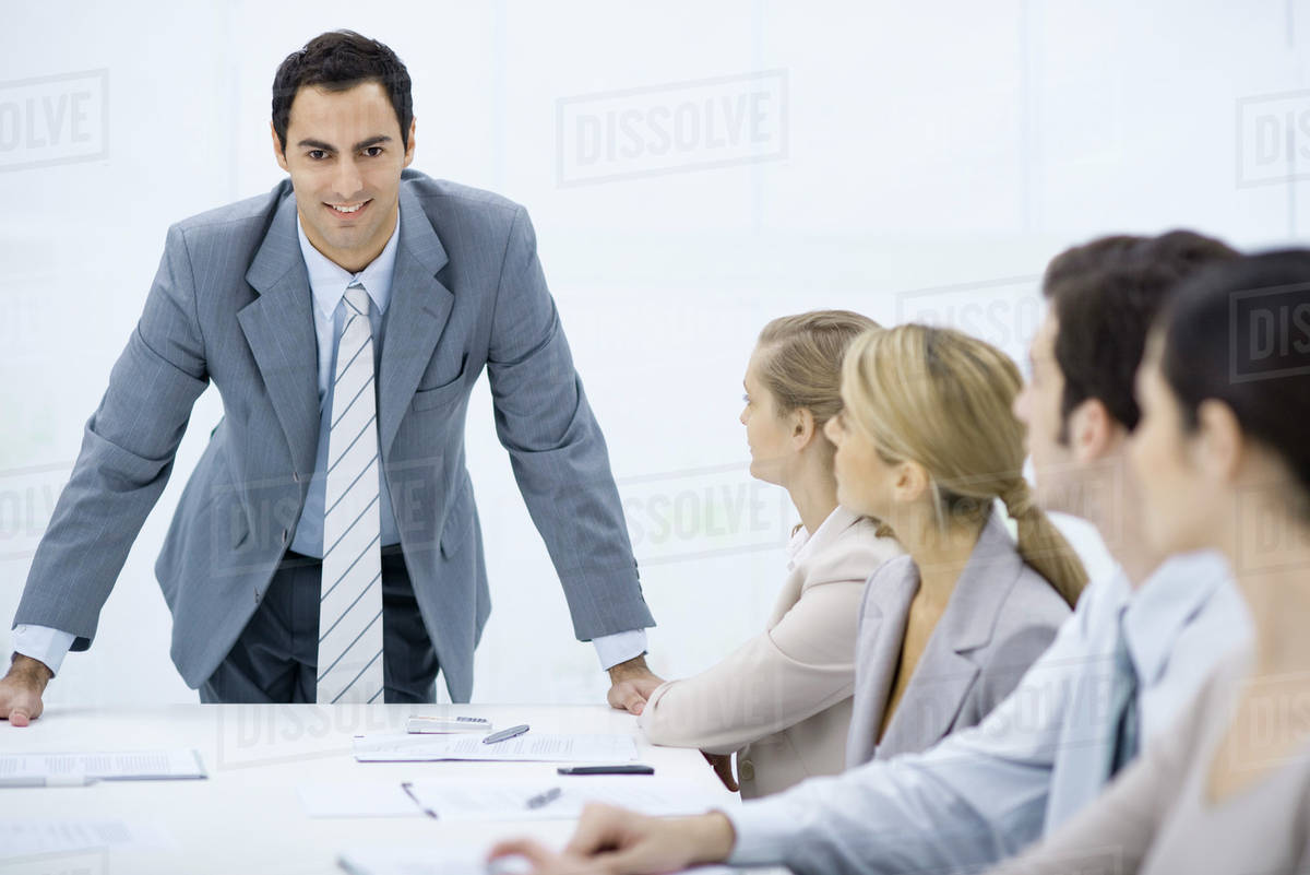 Businessman leaning over table addressing colleagues, smiling at camera ...
