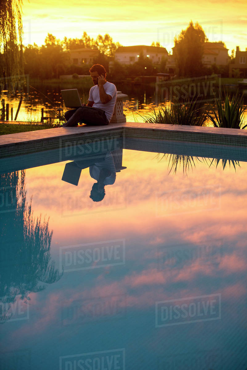 Man using laptop computer next to resort swimming pool as sun sets ...