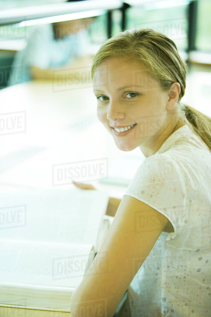 Female college student sitting at table in library, smiling at camera ...