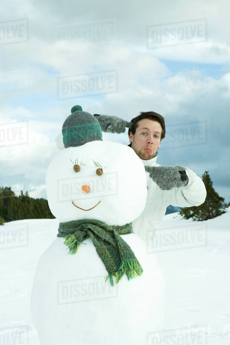 Young man standing behind snowman, pointing, making faces at camera ...
