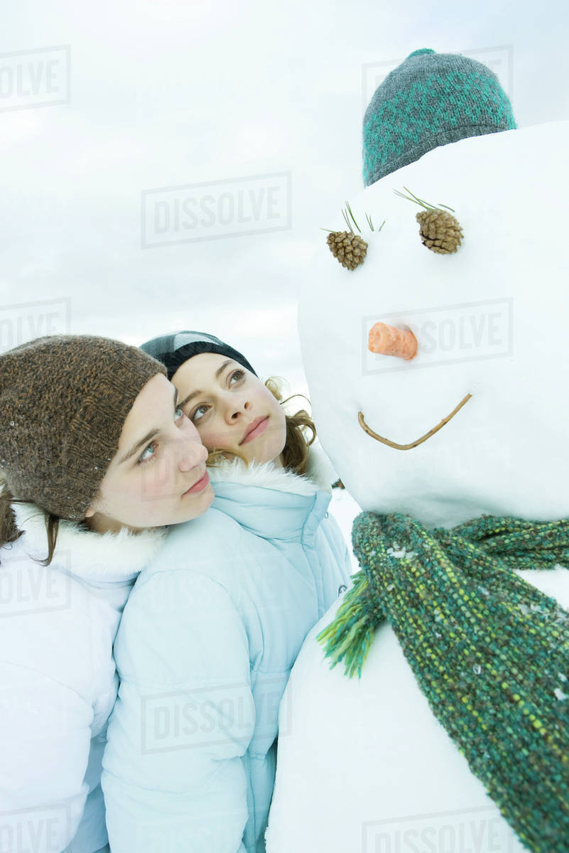 Two young friends looking at snowman together, one leaning on the other ...