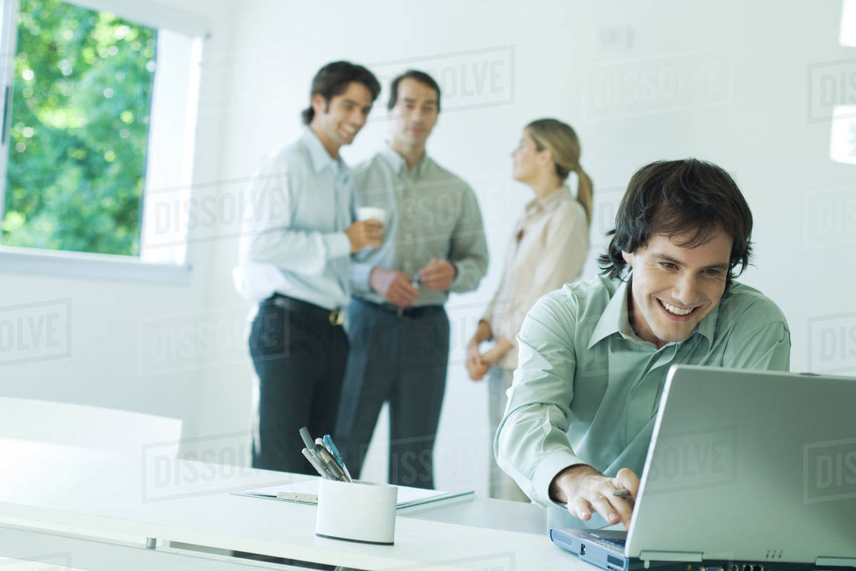 Businessman in office, using laptop computer, smiling, associates in ...