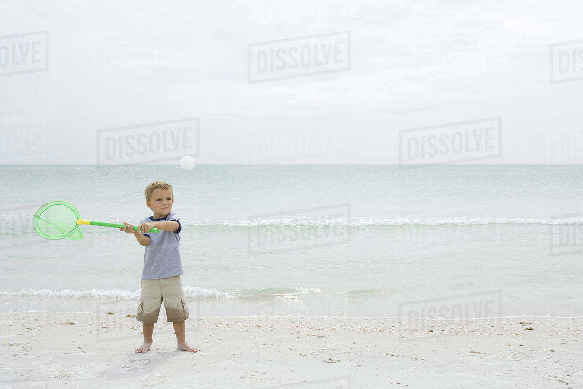 Little boy standing on beach, catching ball with net, full length ...