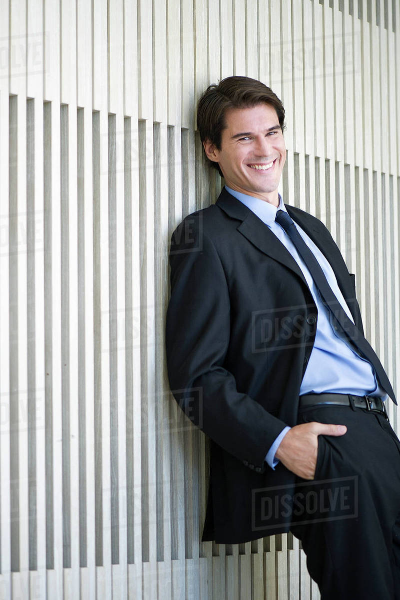 Businessman leaning against wall with hands in pockets, smiling ...