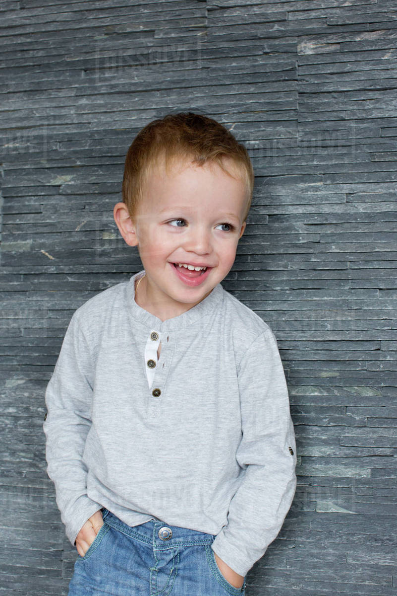 Little boy leaning against wall with hands in pockets, smiling ...