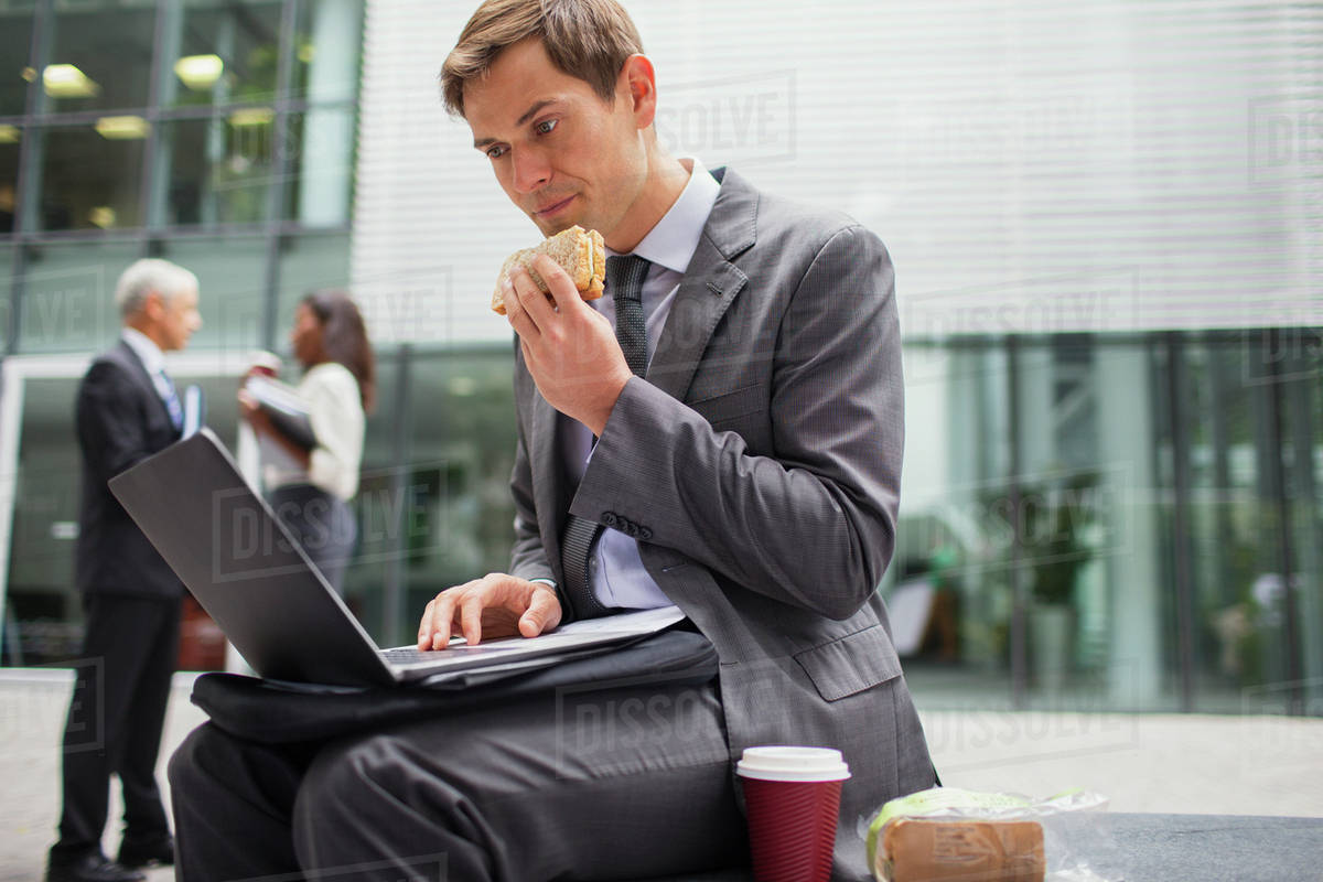 Businessman eating lunch while working outside office building ...