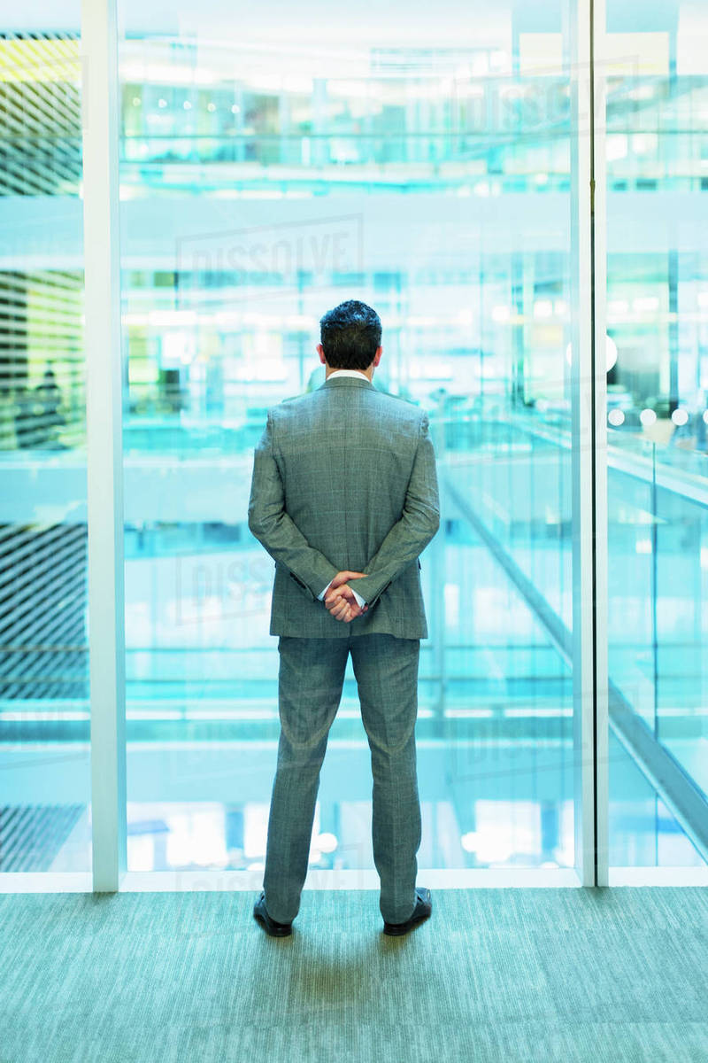 Businessman standing at atrium window with hands behind back - Stock ...