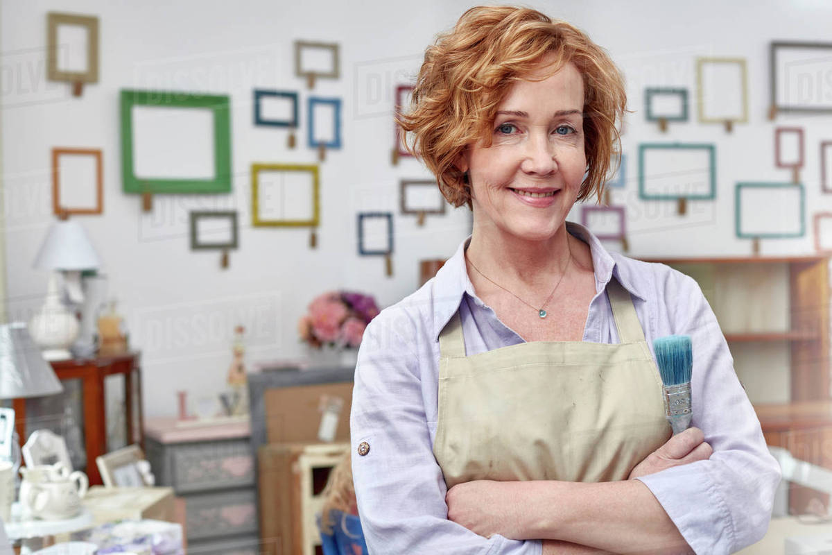 Portrait smiling mature female artist in art shop with picture frames ...