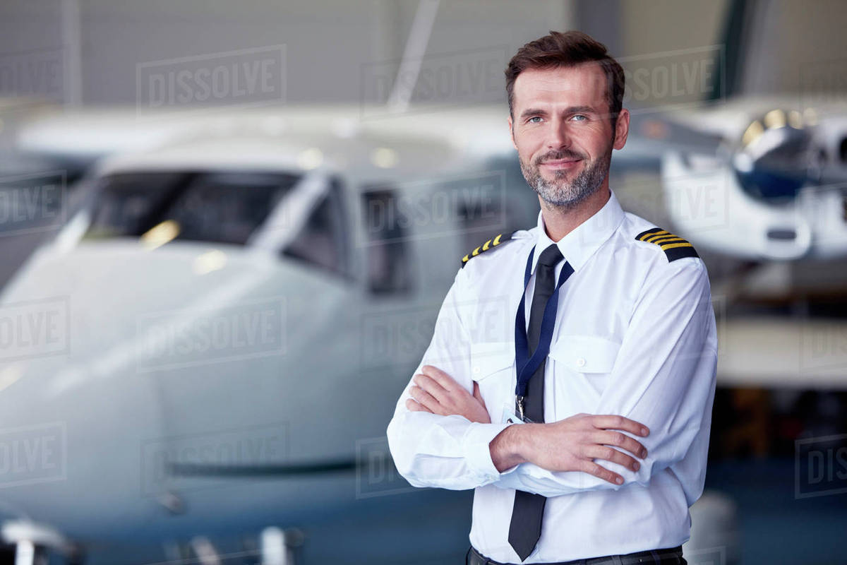 Portrait confident male pilot standing near airplane in hangar ...