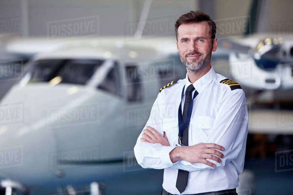 Portrait confident male pilot standing near airplane in hangar ...
