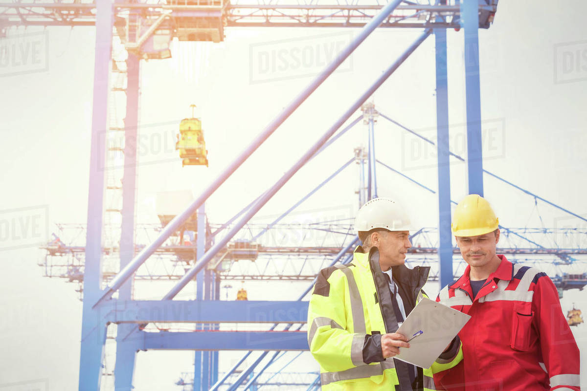 Dock worker and manager with clipboard talking below crane at shipyard ...