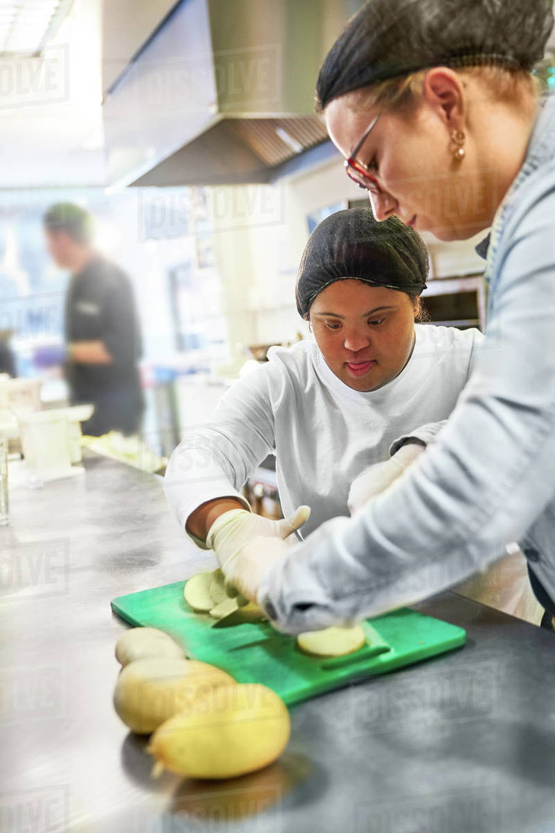 Chef and young woman with Down Syndrome cutting potatoes in kitchen ...