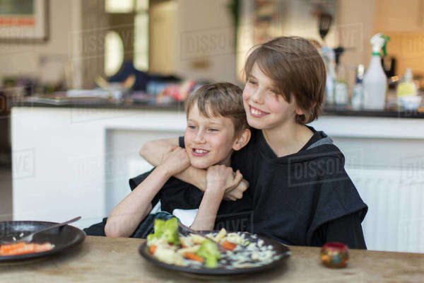Happy brothers hugging and eating dinner at dining table - Royalty-free ...