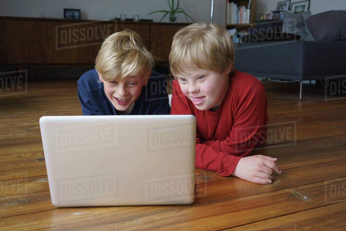 Smiling brothers using laptop while lying on hardwood floor at home ...