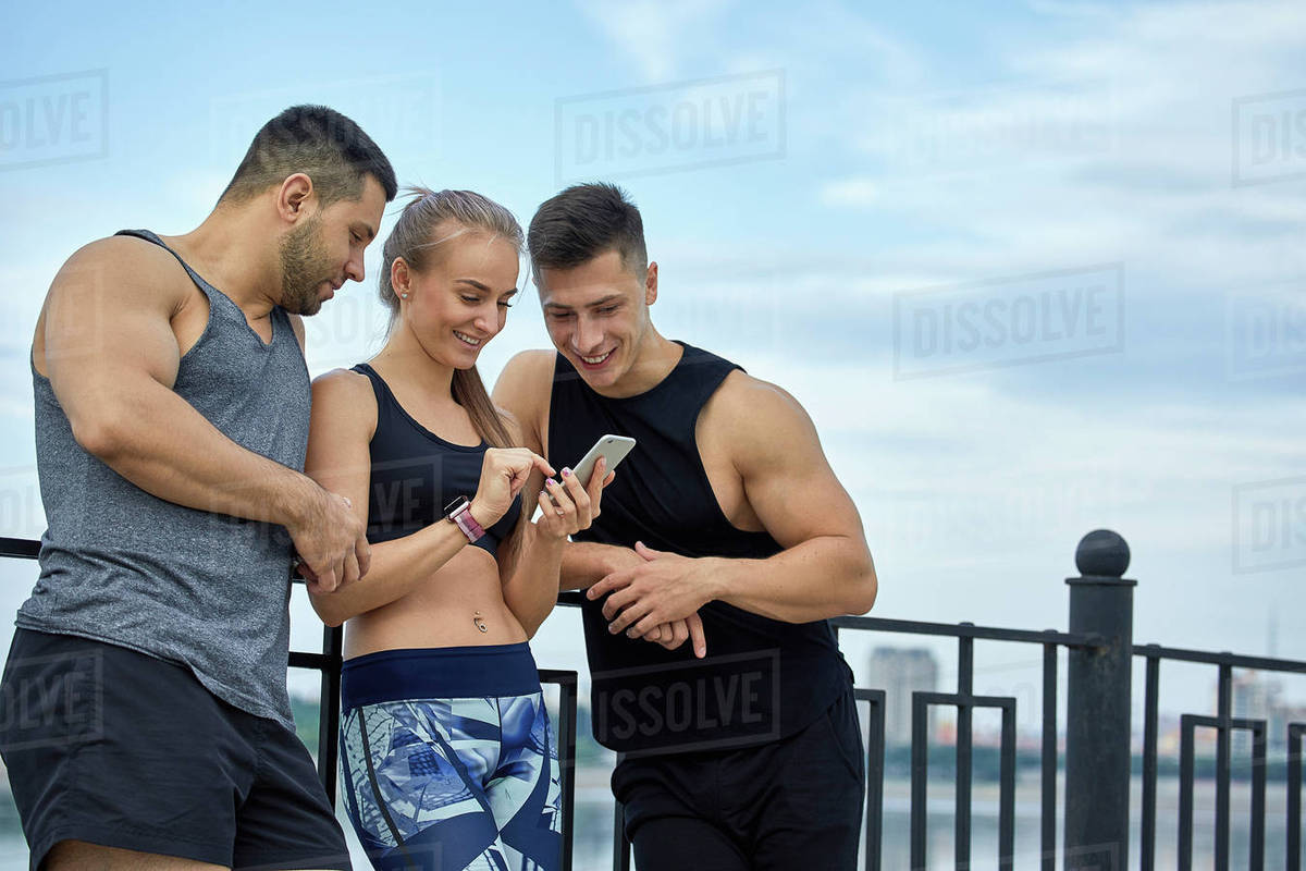 Happy young athletes using mobile phone while leaning by railing ...