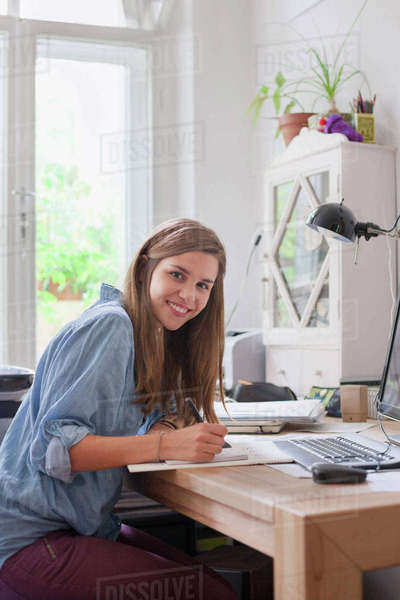 Portrait of smiling young woman writing at computer table in room ...