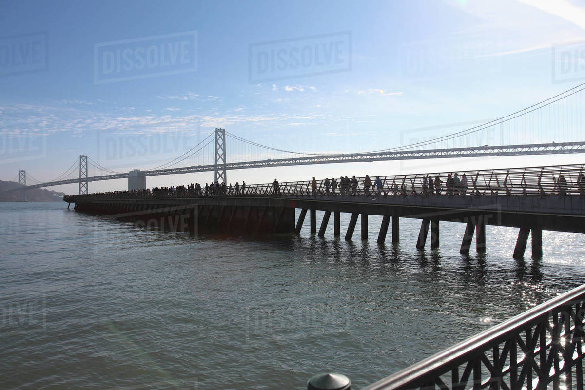 People on pier with Oakland Bay Bridge over bay in background, San ...
