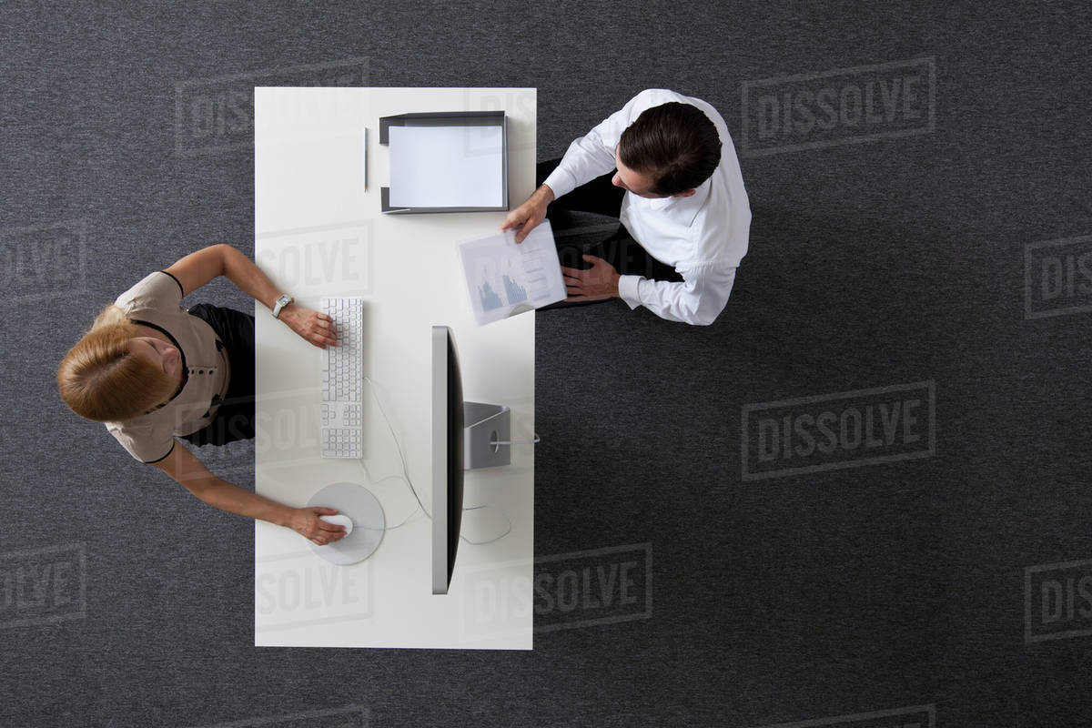A businessman giving papers to a businesswoman sitting at a desk ...